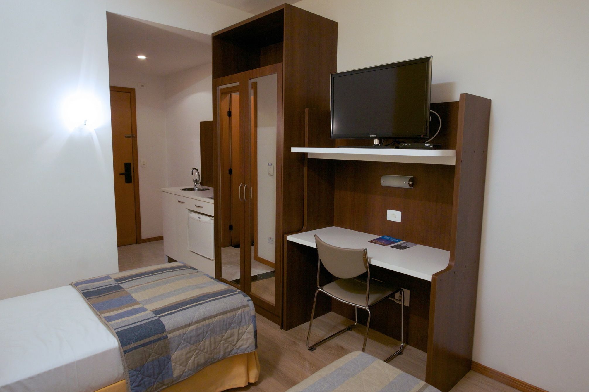 Quarto compacto com cama, escrivaninha, cadeira, televisão e uma pequena cozinha equipada.