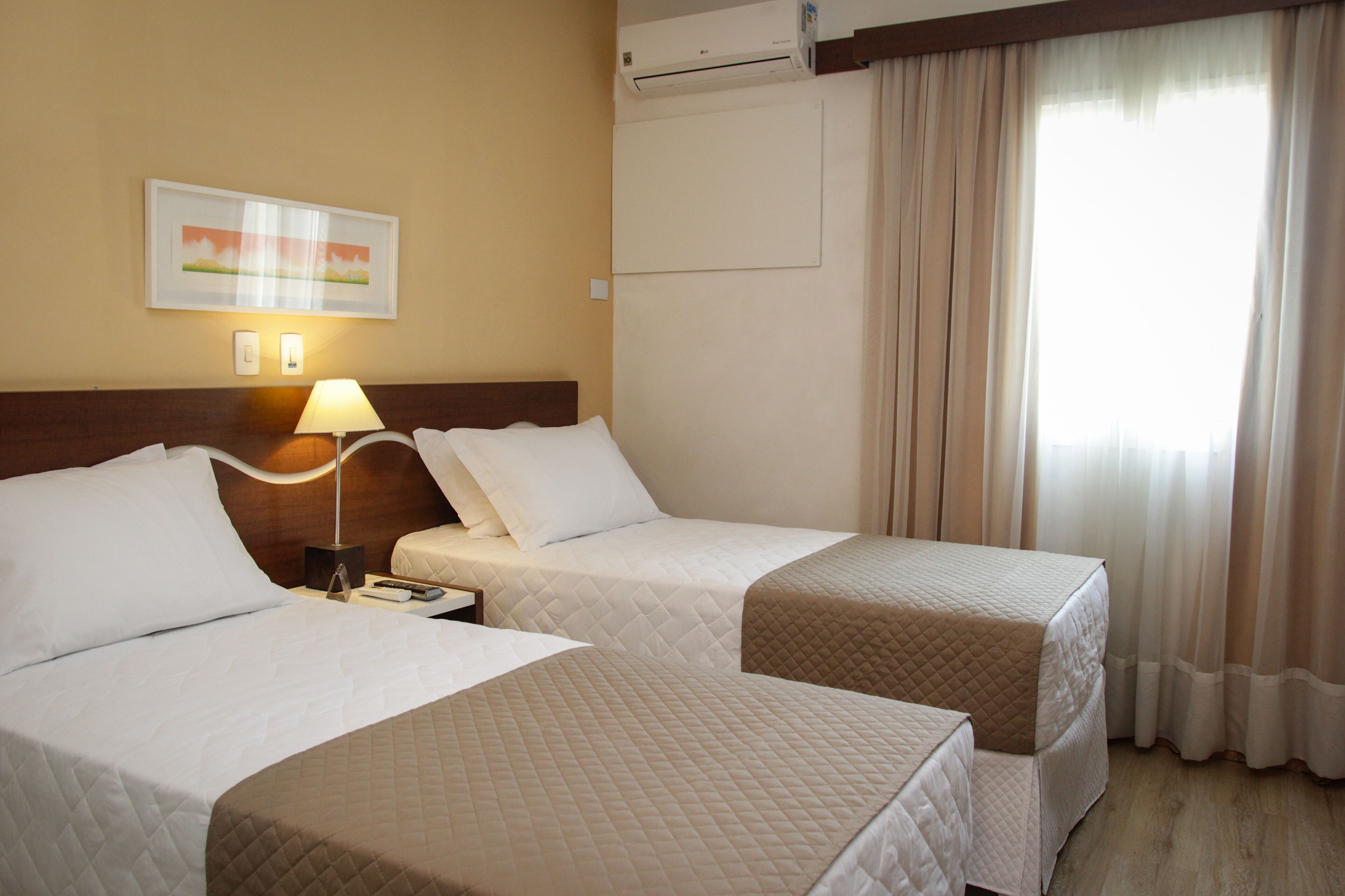 Quarto de hotel com duas camas, uma mesa de cabeceira com abajur, aparelho de ar condicionado e janela com cortinas.