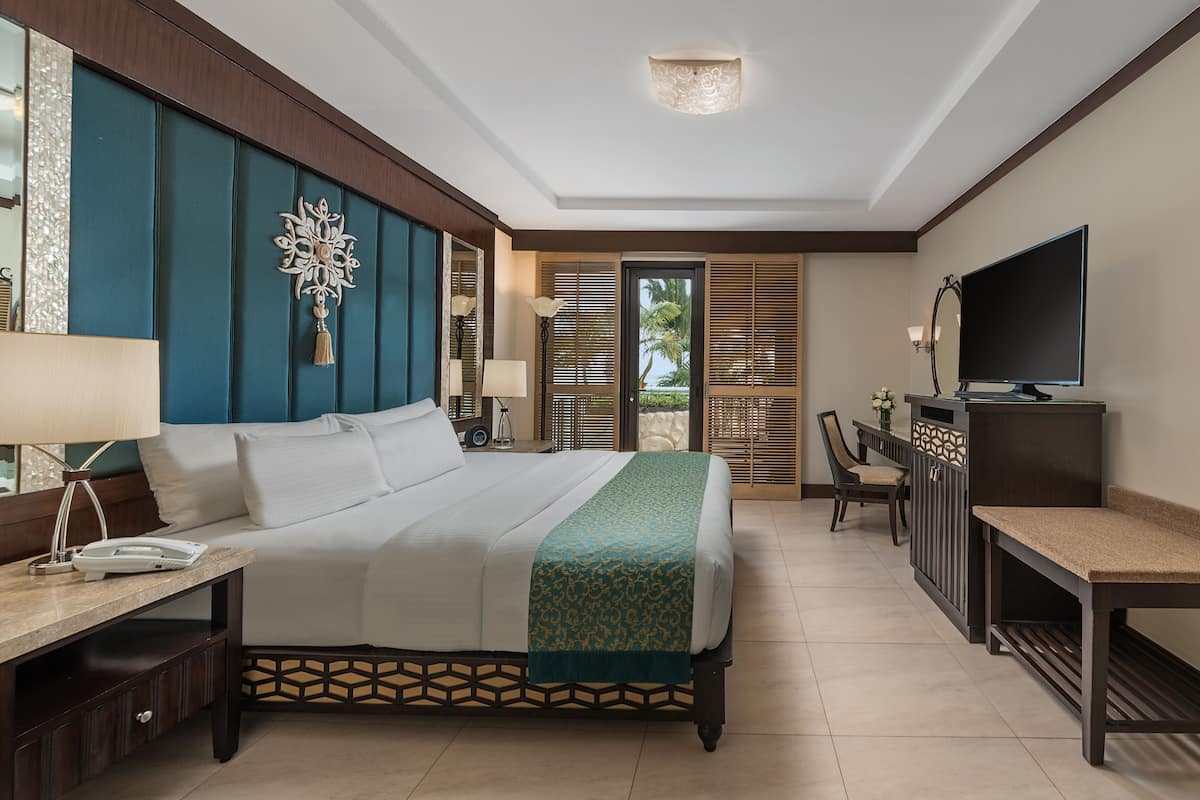 Quarto de hotel com uma cama grande, uma mesa com luminária, uma televisão e vista para o exterior através de uma porta.