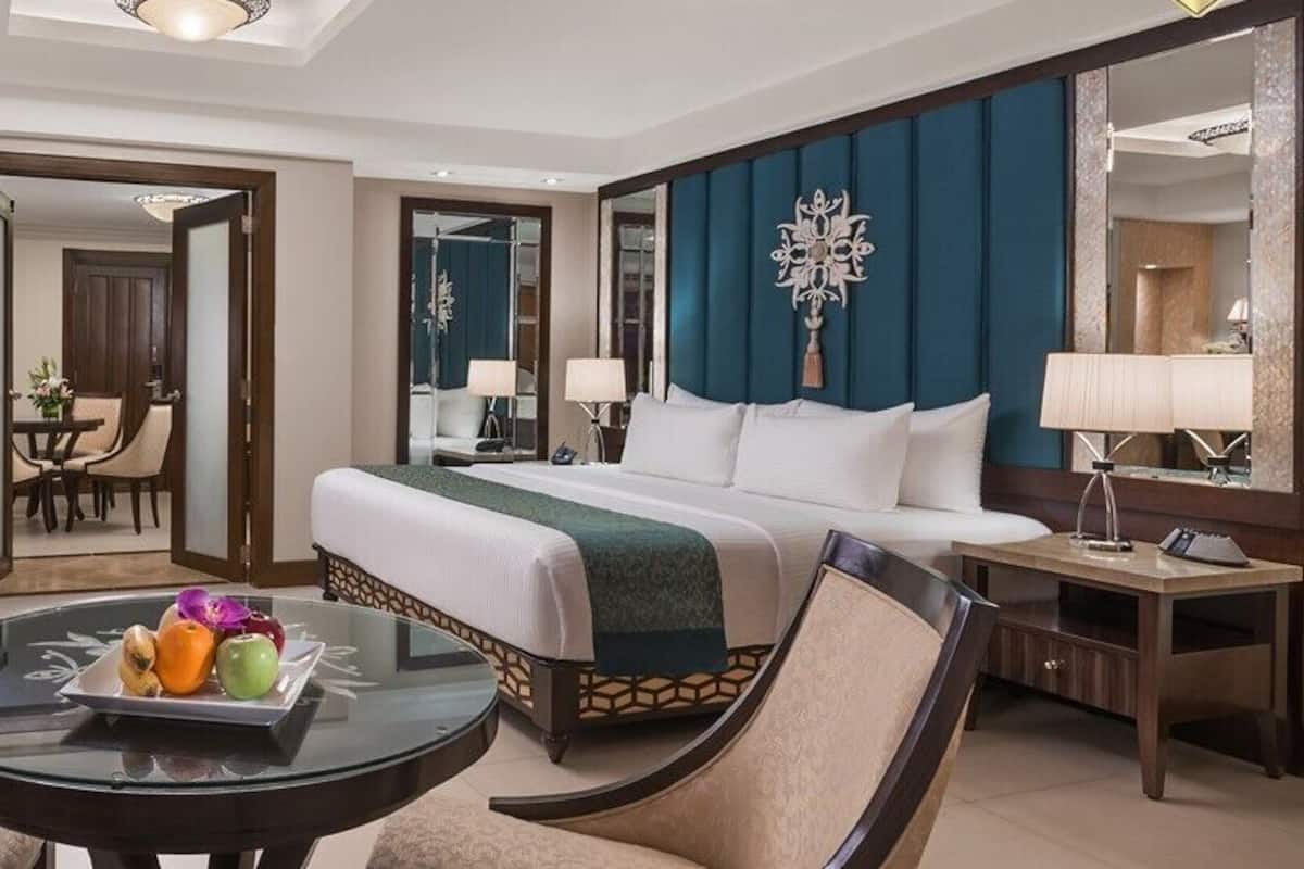 Quarto de hotel com cama, escrivaninha, cadeira, mesa redonda com frutas e televisão.