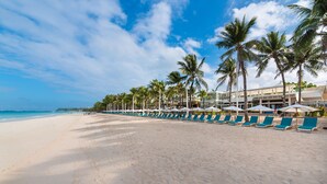Uma praia com fileiras de espreguiçadeiras e guarda-sóis, palmeiras e um edifício com design moderno.