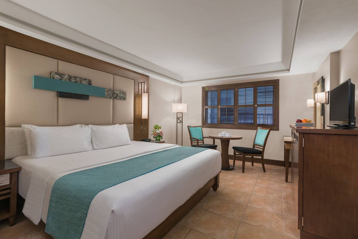 Quarto de hotel com uma cama grande, uma escrivaninha com cadeira, uma mesinha e uma televisão.