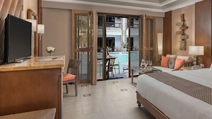 Quarto de hotel com uma cama grande, uma mesa com TV, área de refeições com mesa e cadeiras, e vista para uma piscina e um edifício.