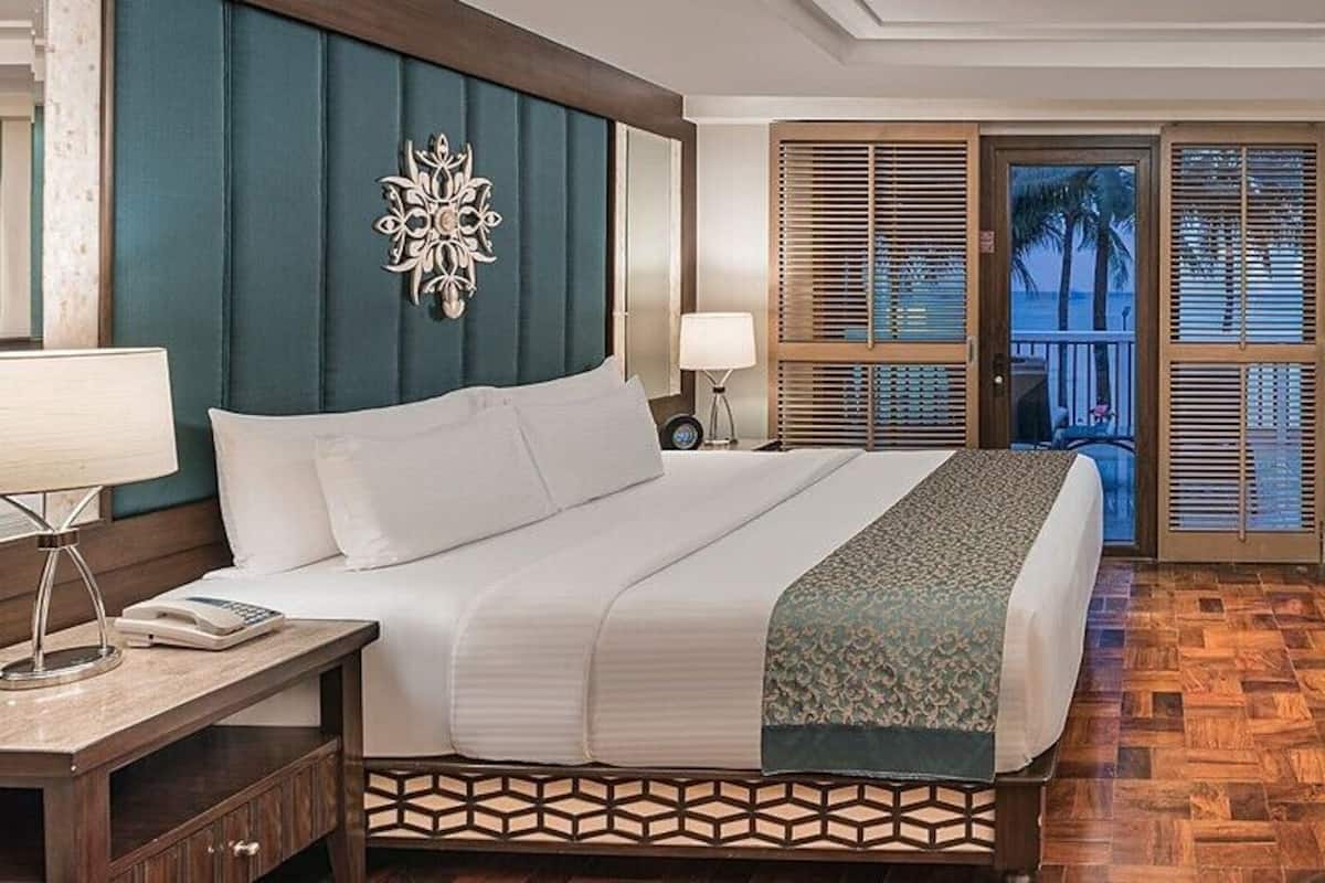 Quarto de hotel com cama, mesa de cabeceira, luminária e vista para a praia pela janela.