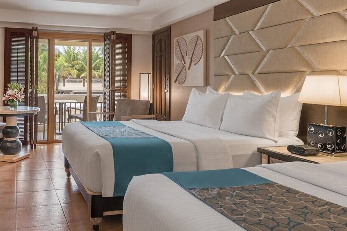 Quarto de hotel com duas camas, área de refeições e vista para palmeiras.