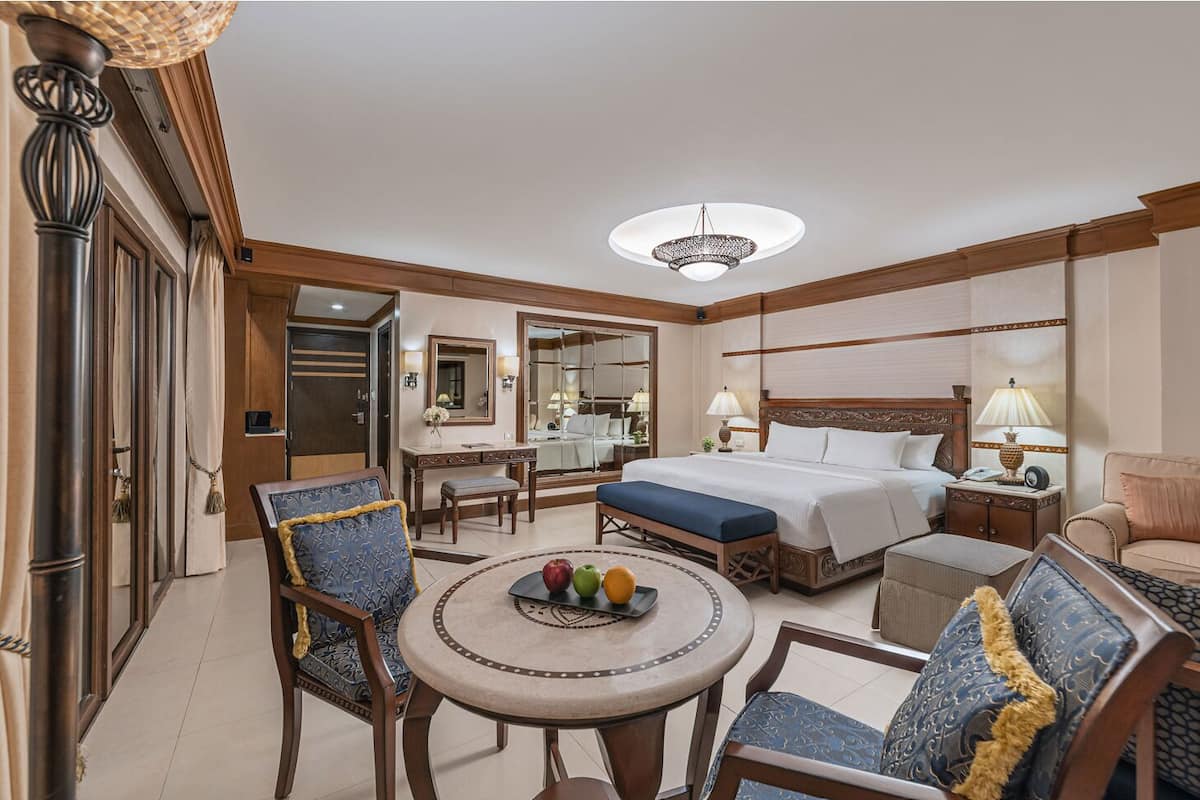 Quarto de hotel com cama, escrivaninha, cadeira, mesa redonda com frutas e um espelho grande.