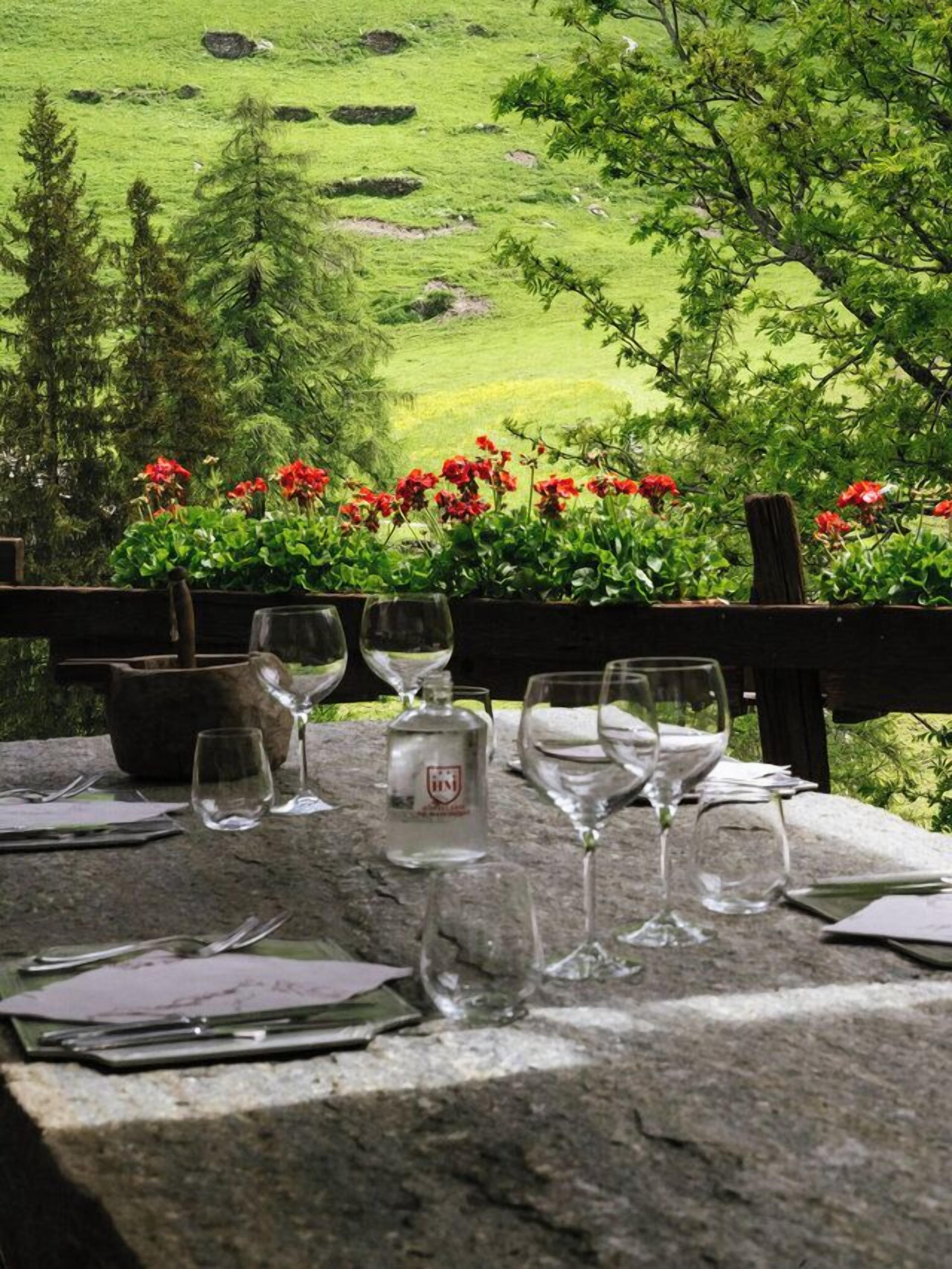 Mesa posta ao ar livre com copos de vinho, garrafa e jarra, com vista para um campo verde com flores vermelhas.