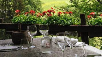 Mesa posta ao ar livre com copos de vinho, garrafa e jarra, com vista para um campo verde com flores vermelhas.