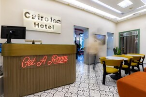 Uma área de recepção de hotel com um letreiro 'Get A Room', um balcão de atendimento e uma área de estar com cadeiras e mesas amarelas.