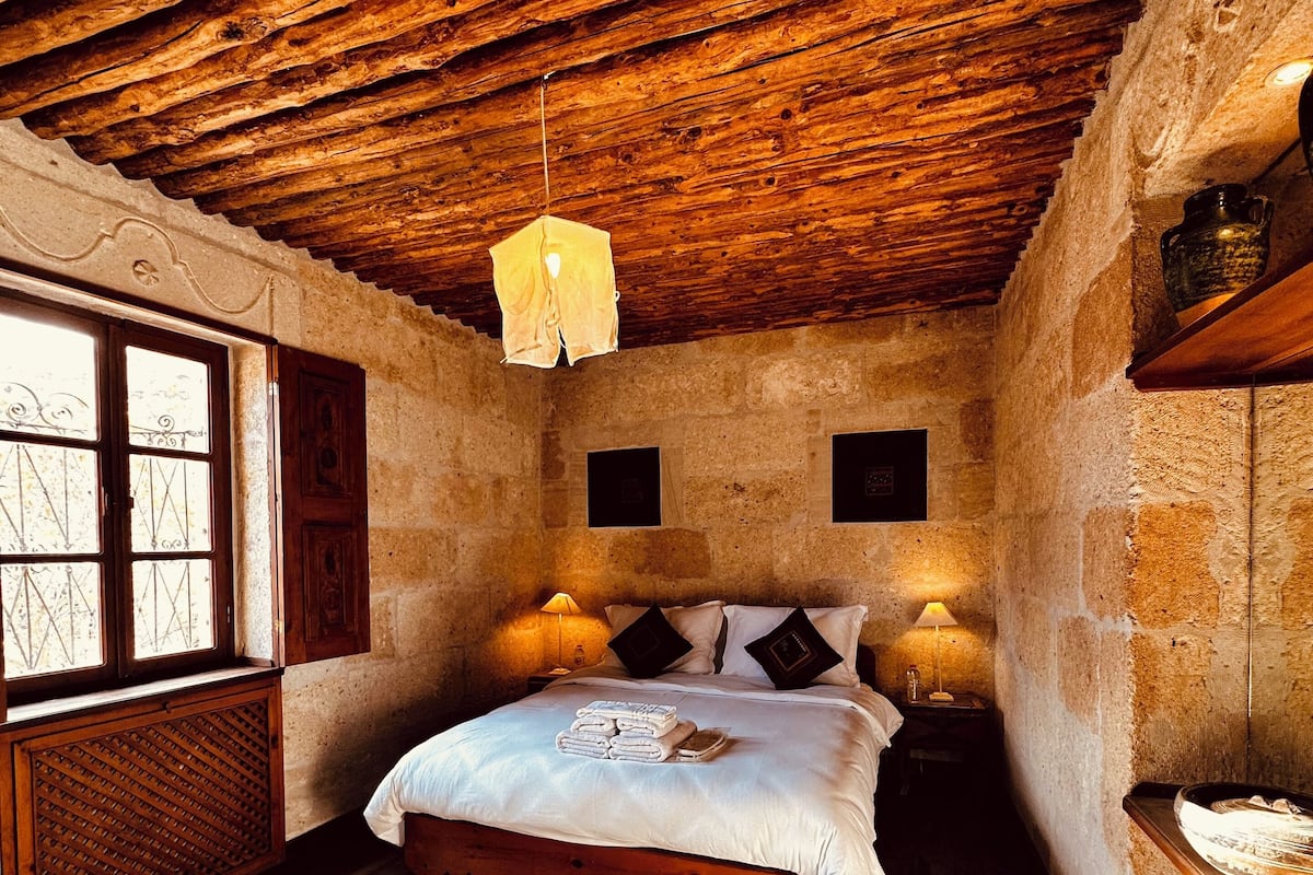 Quarto rústico com teto de madeira, cama com lençóis brancos, cabeceira de madeira, janela com grades e uma pequena mesa com abajur.