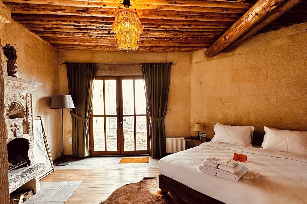 Quarto rústico com teto de madeira, cama com lençóis brancos, lareira e vista através de uma grande janela.