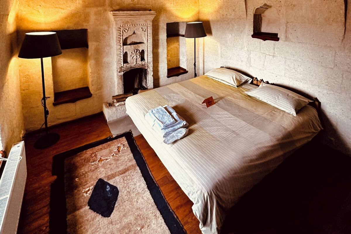 Quarto com teto de pedra grande e arqueado, cama com colcha branca, duas luminárias de cabeceira, lareira e uma pequena mesa com um abajur.