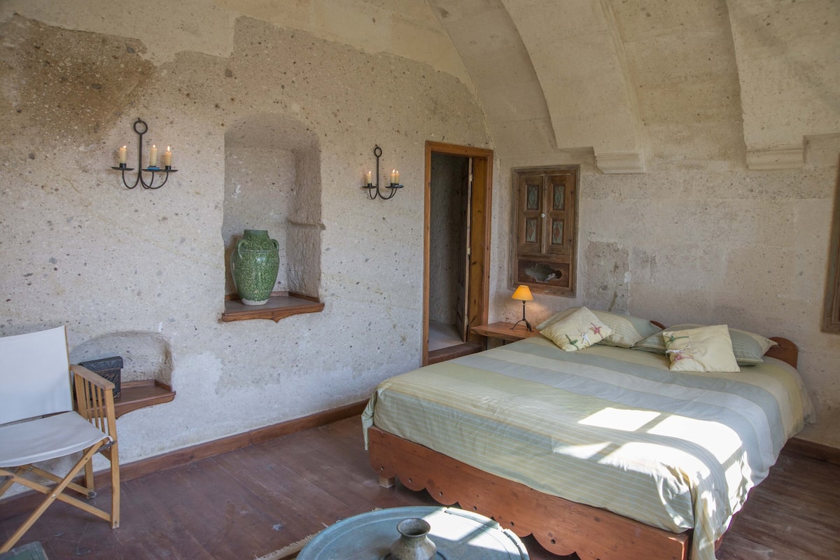 Quarto rústico com cama de madeira, uma mesinha, uma cadeira e um vaso verde numa prateleira.