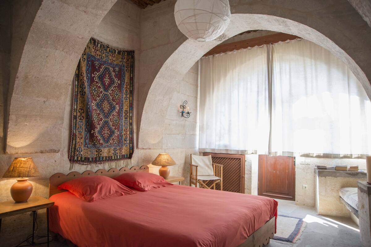 Um quarto com cama, duas luminárias, uma cadeira, tapeçaria e janela com cortinas.