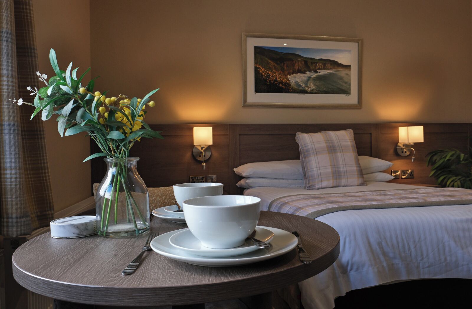 Quarto de hotel com cama, mesas de cabeceira, mesa de jantar com tigelas e um vaso de flores, e um quadro na parede.