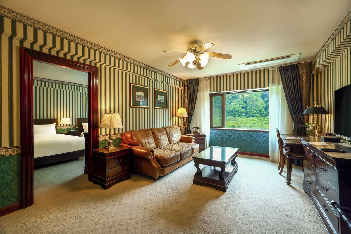 Quarto de hotel com cama, sofá de couro, mesa de centro, escrivaninha e vista para o verde lá fora.