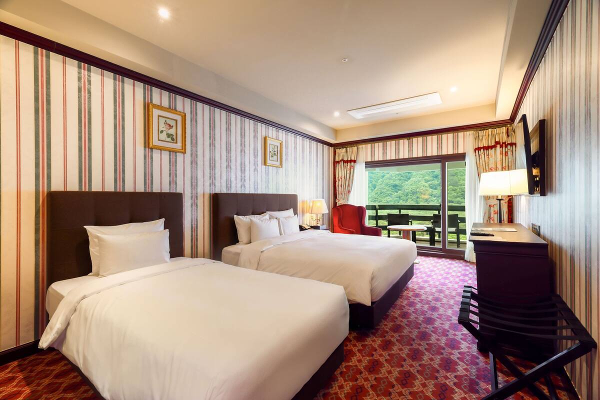 Quarto de hotel com duas camas, uma escrivaninha, uma cadeira e vista para o exterior.
