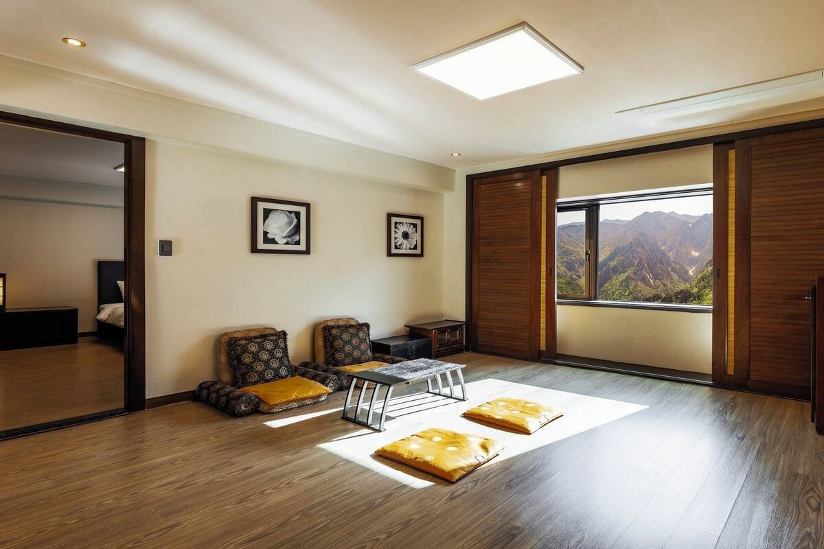 Uma sala de estar espaçosa com uma janela ampla que oferece vista para as montanhas, um conjunto de sofás e uma mesinha.