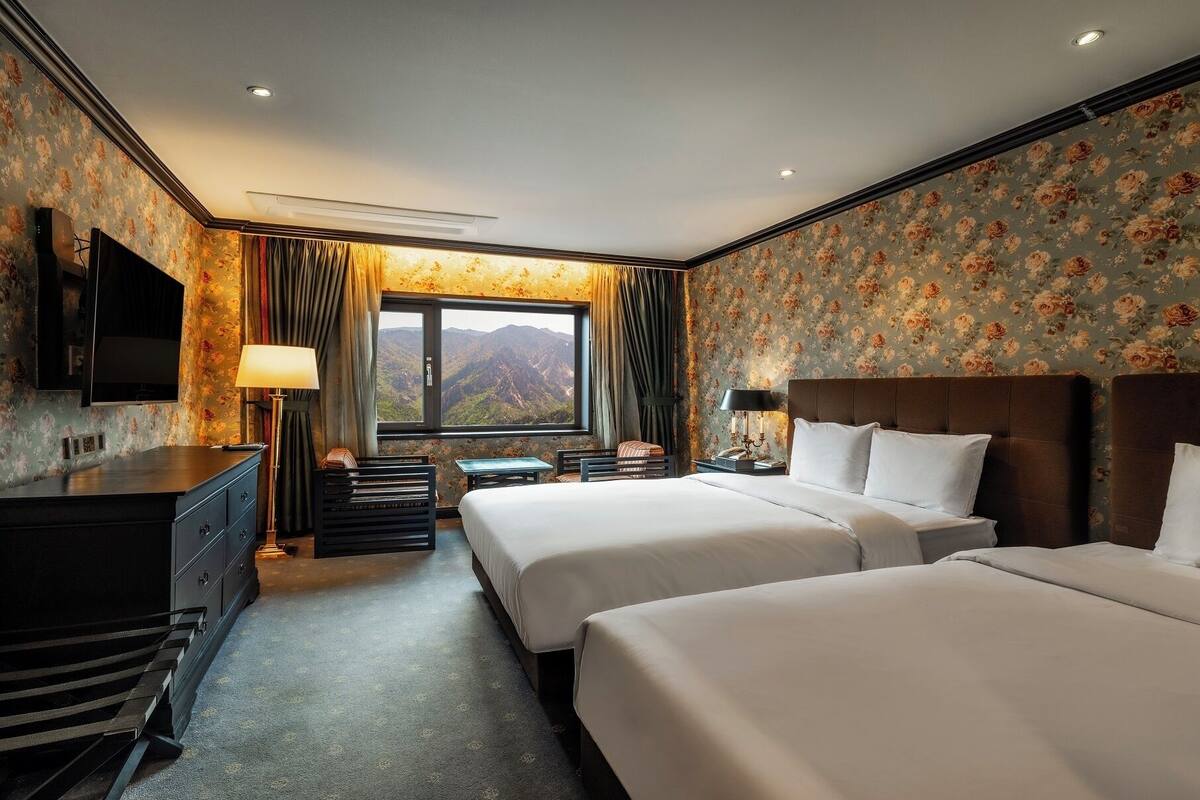 Quarto de hotel com duas camas, uma escrivaninha, televisão e janela com vista para as montanhas.