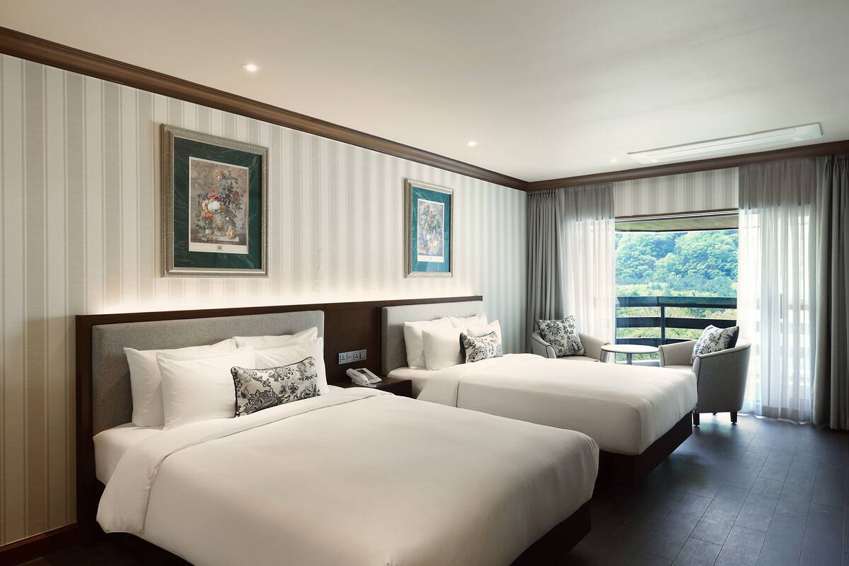 Quarto de hotel com duas camas, uma mesinha, uma cadeira e varanda com cortinas.