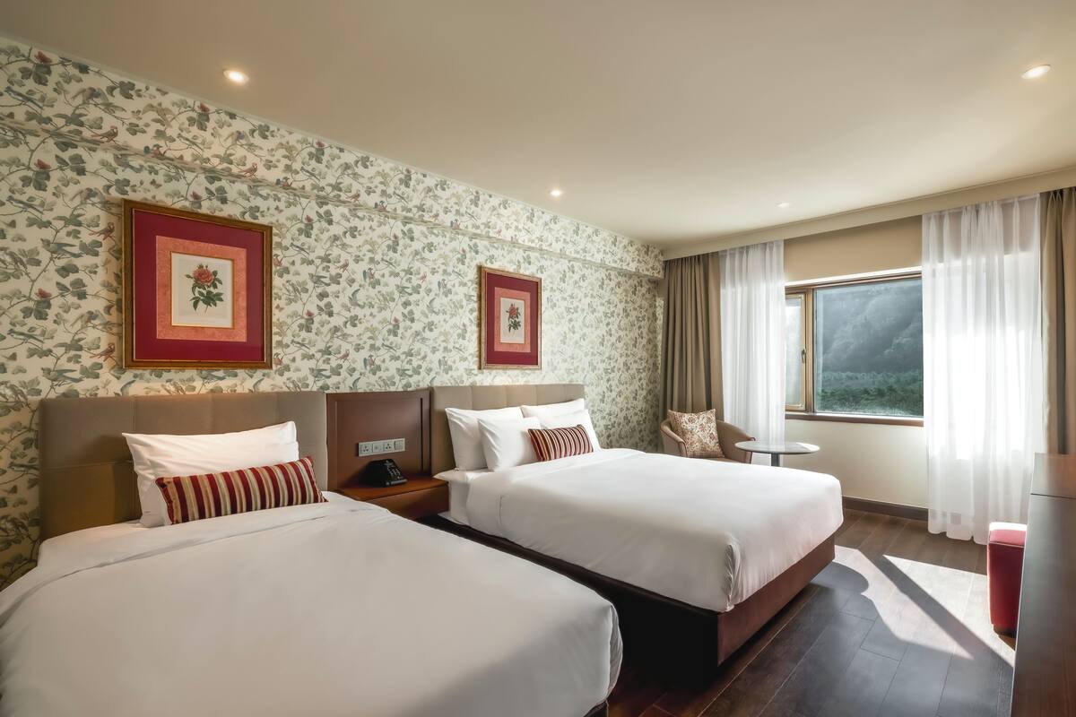 Quarto de hotel com duas camas, papel de parede floral, quadros emoldurados, uma mesa pequena e uma janela com cortinas.
