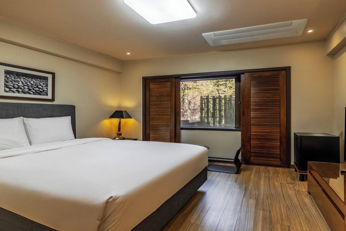 Quarto de hotel com uma cama, mesa de cabeceira com abajur, televisão e janela com persianas de madeira.