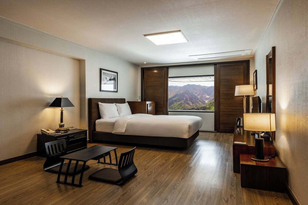 Quarto de hotel com cama, mesa de cabeceira, luminária, escrivaninha, cadeira e vista para as montanhas.