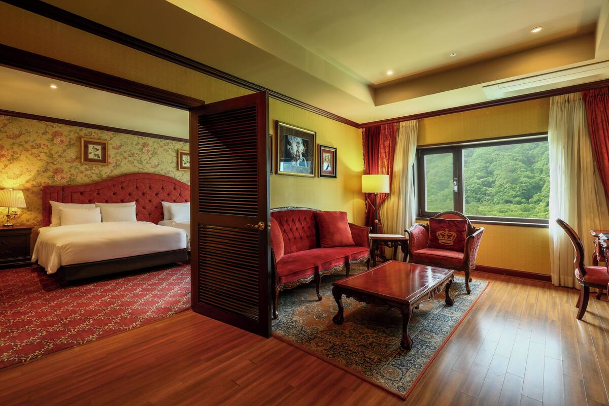 Quarto de hotel com cama, sofá vermelho, mesa de centro de madeira e vista para o verde pela janela.