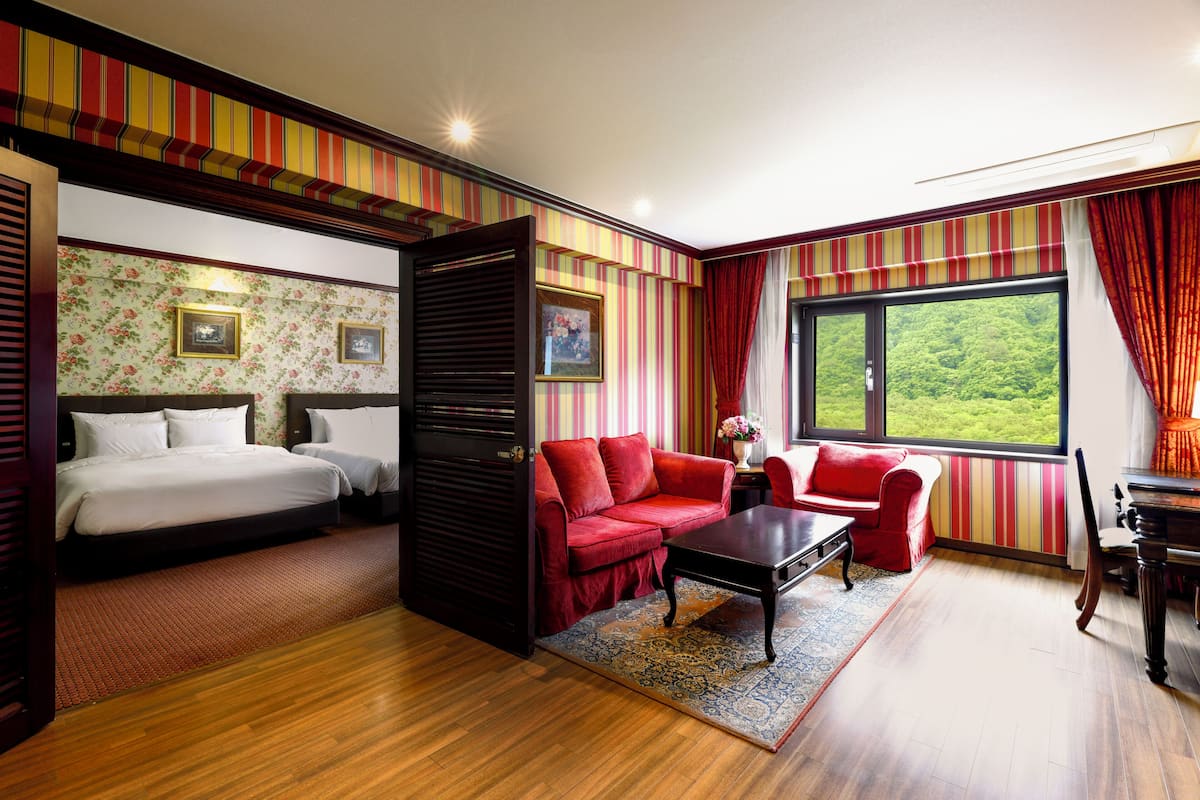 Quarto com cama, sofá vermelho, mesa de centro e janela com vista para vegetação.