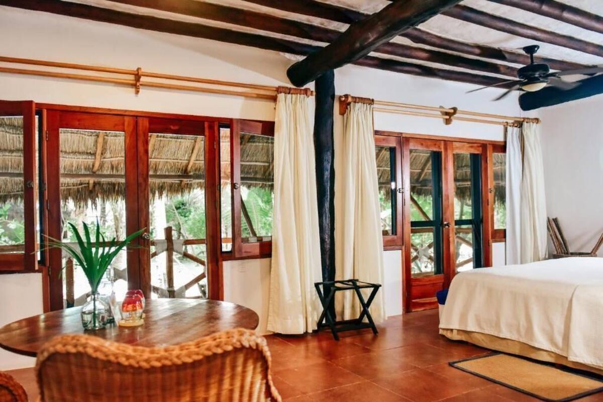 Quarto com cama, mesa e cadeira. Janelas com cortinas e ventilador no teto.