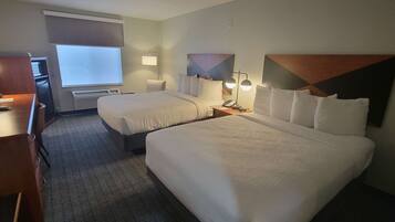 Quarto de hotel com duas camas, uma escrivaninha, uma cadeira, um abajur e uma janela.