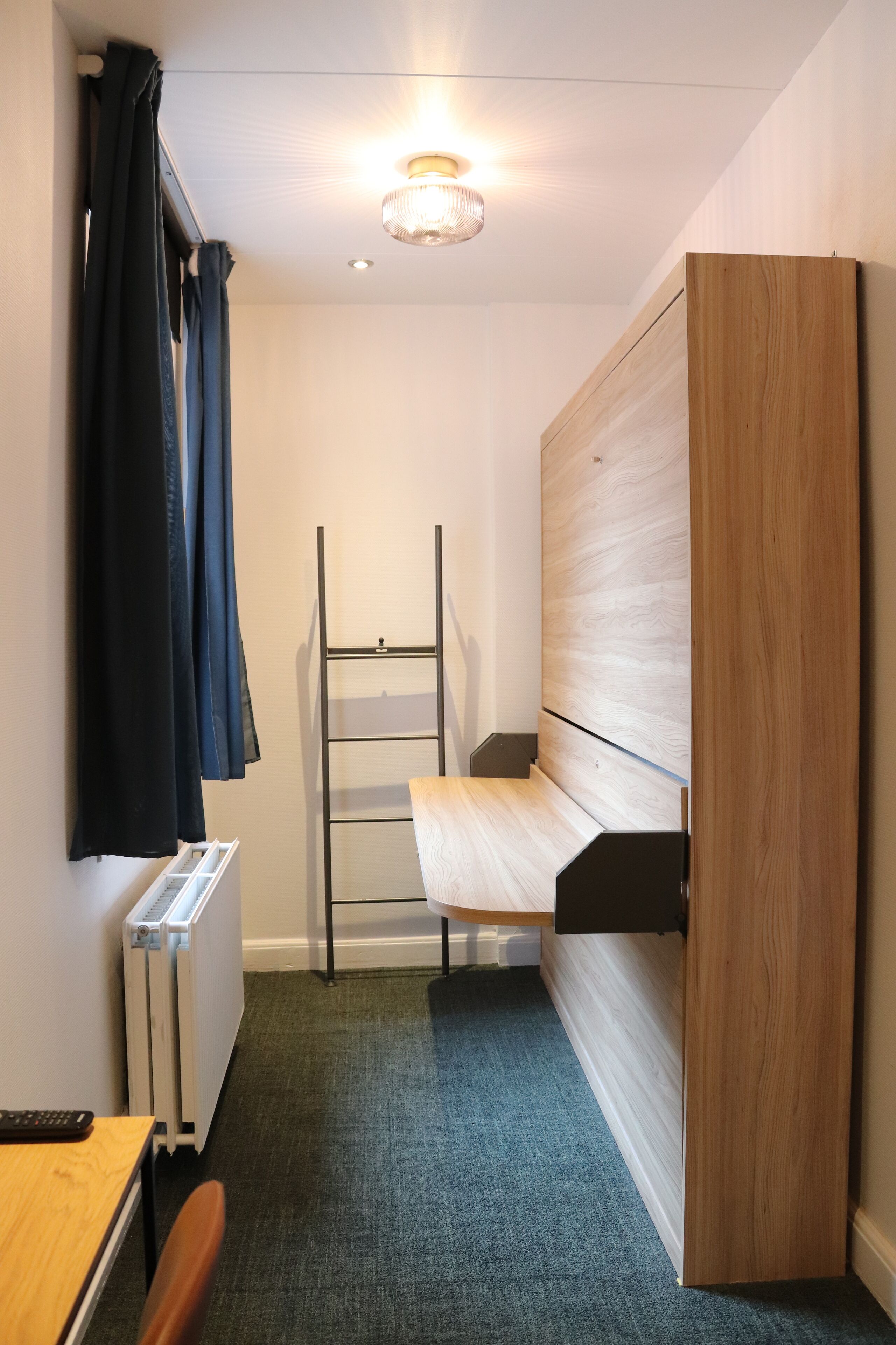 Quarto pequeno e compacto com uma cama beliche, um radiador, uma escrivaninha e uma cadeira.