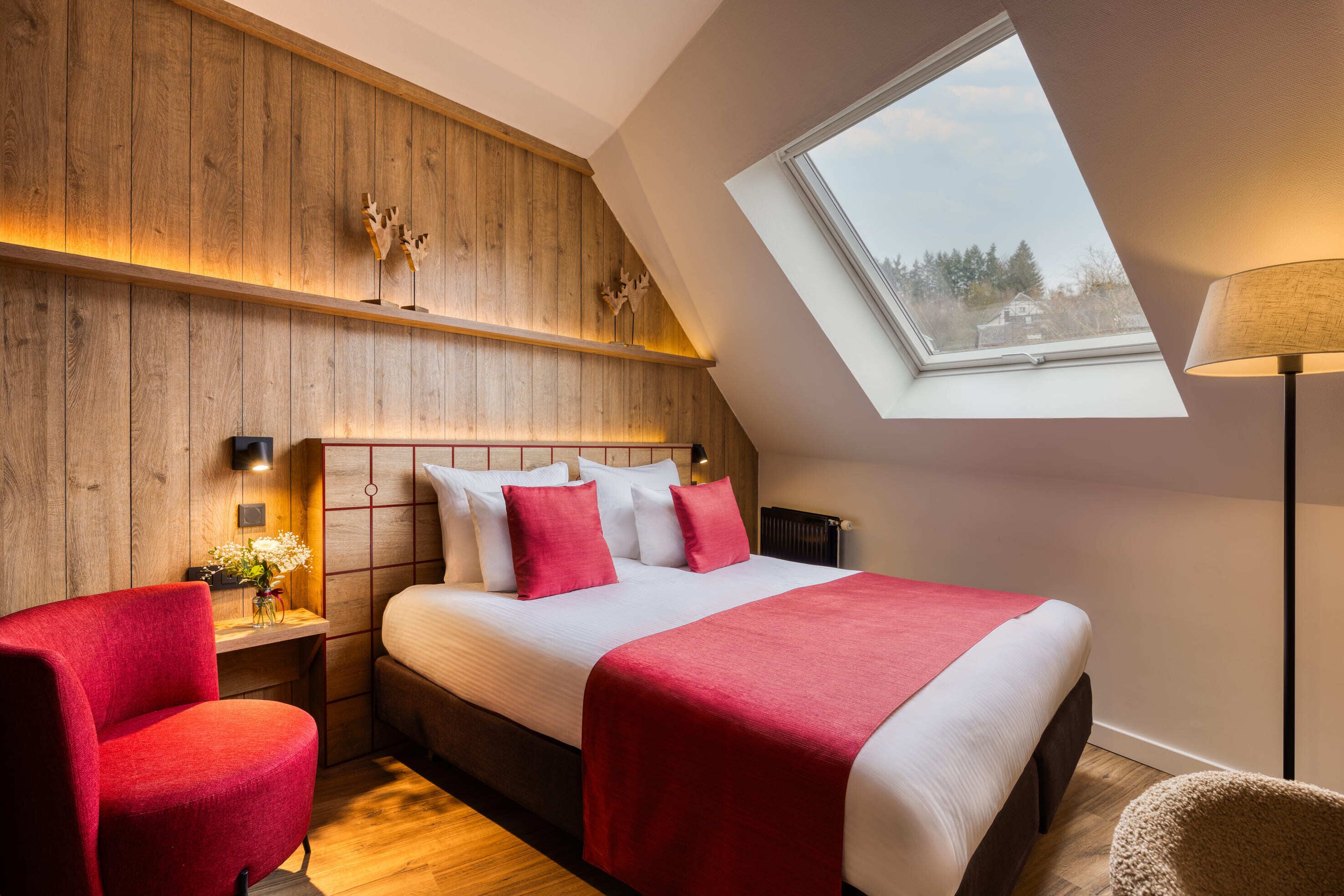 Quarto com teto inclinado, claraboia, cama com roupa de cama branca e vermelha, poltrona vermelha e mesa de cabeceira com abajur.
