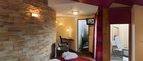 Quarto de hotel com parede de pedra, cama com manta vermelha, televisão na parede e varanda com cadeiras.