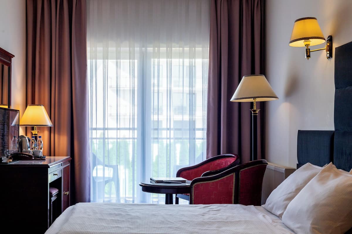 Quarto de hotel com uma cama, duas luminárias de cabeceira, uma escrivaninha com abajur, uma cadeira e vista através de cortinas leves.