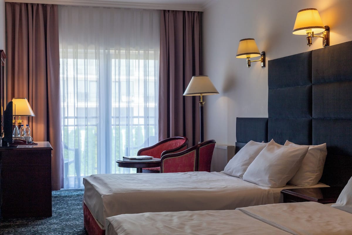 Quarto de hotel com duas camas, uma escrivaninha com luminária, uma cadeira e uma janela grande com cortinas.