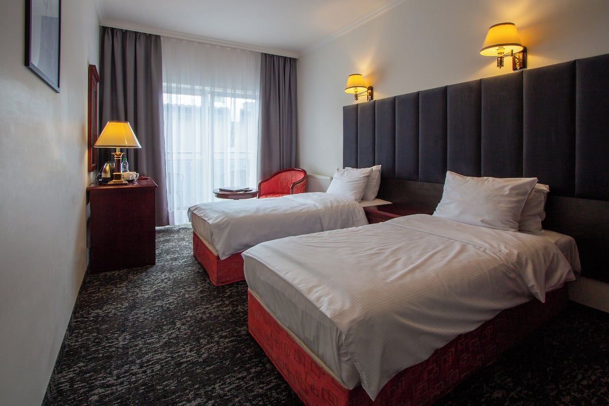 Quarto de hotel com duas camas, mesa de cabeceira com abajur, uma cadeira vermelha e janela com cortinas.