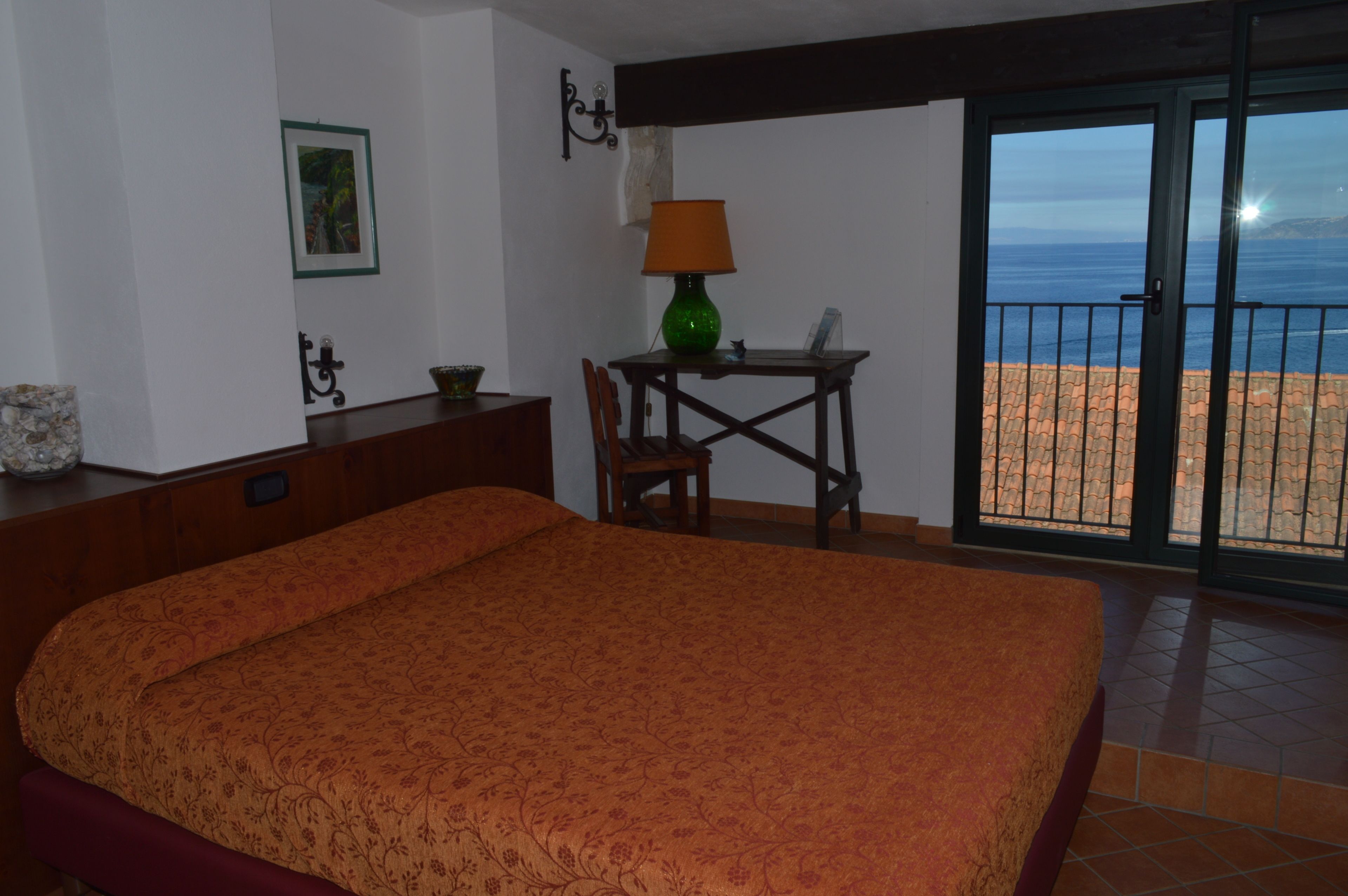 Quarto com cama, escrivaninha iluminada por um abajur, cadeira e vista para a praia através de uma porta de correr.