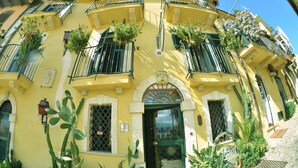 Um edifício amarelo com porta verde, várias varandas com plantas e uma rua de calçada.