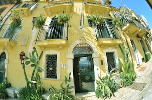 Um edifício amarelo com porta verde, várias varandas com plantas e uma rua de calçada.