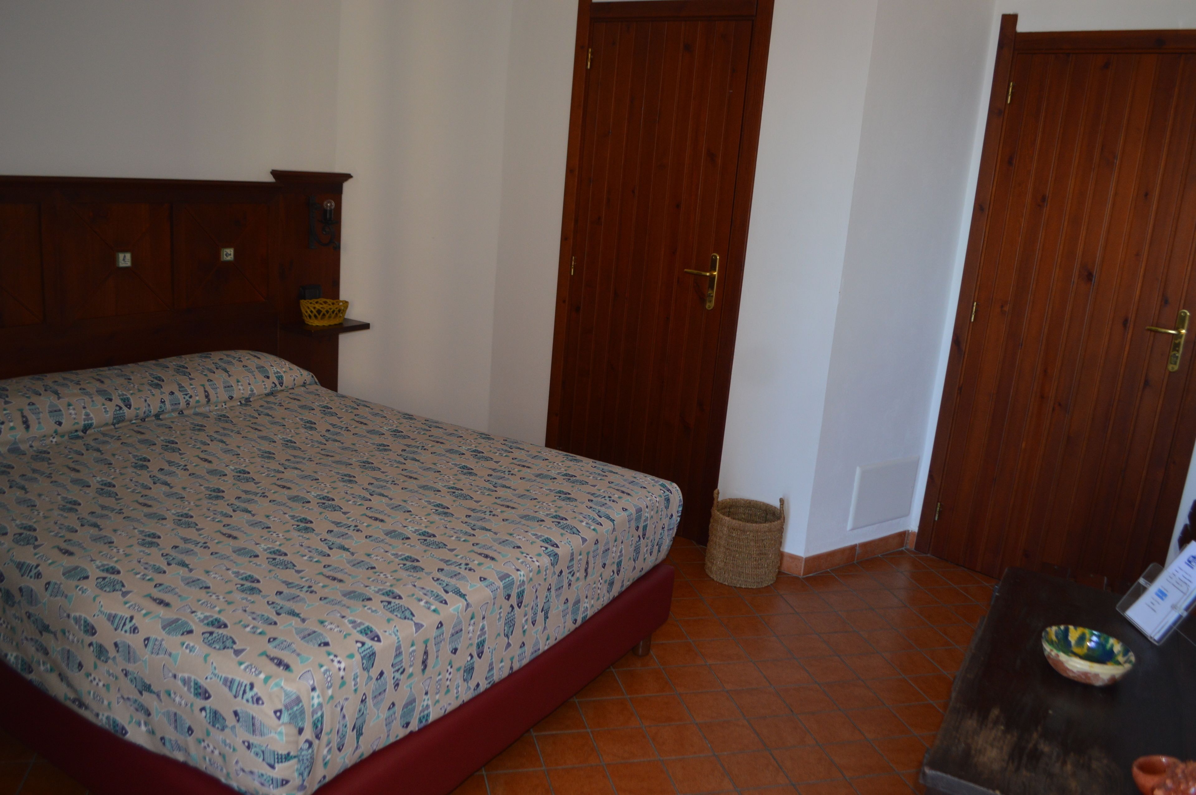 Quarto com cama de madeira, duas portas de madeira, uma mesa pequena com uma tigela e uma cesta de vime no chão.