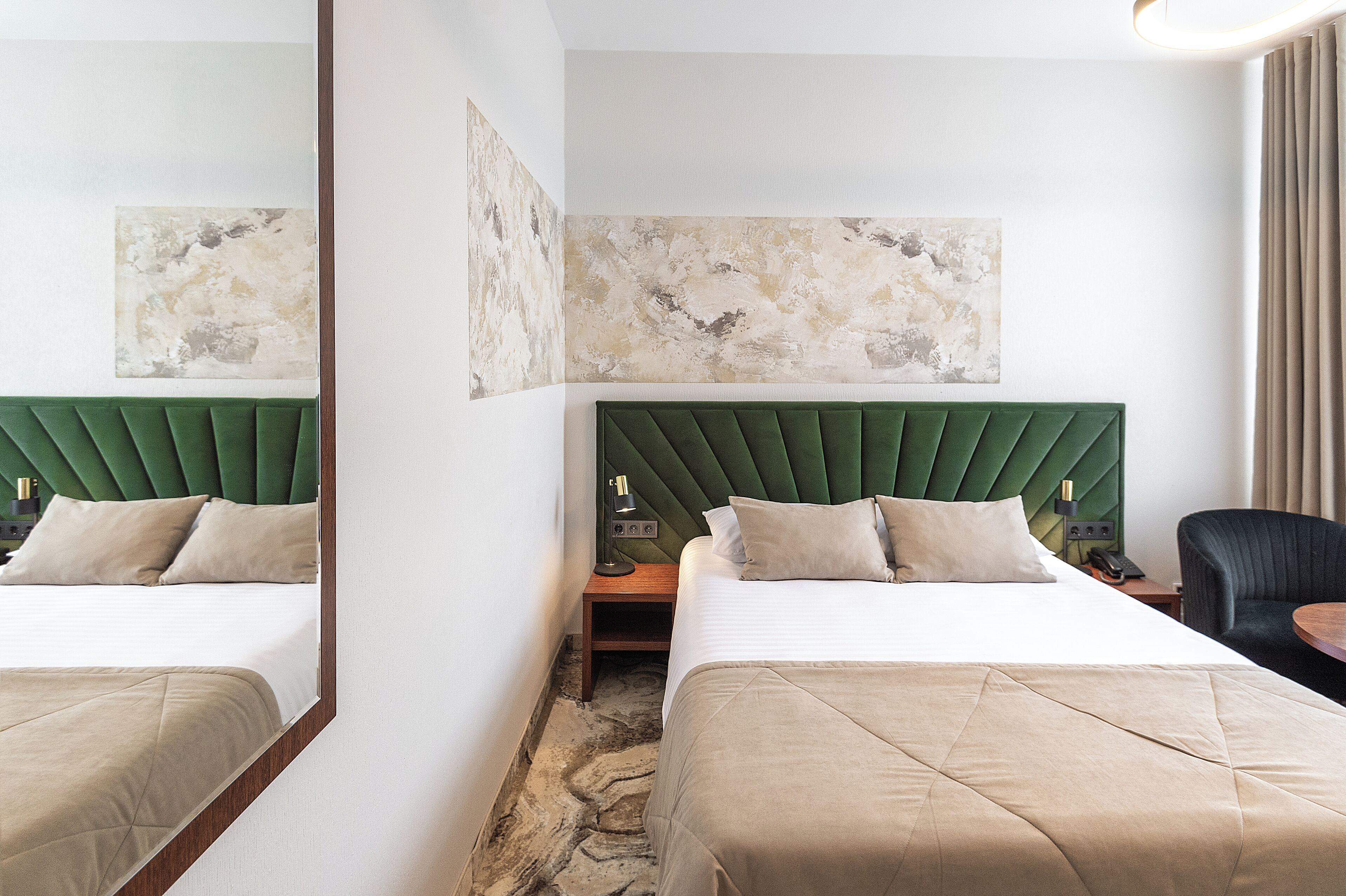 Quarto de hotel com uma cama grande, cabeceira estofada verde, mesa de cabeceira com abajur e uma obra de arte na parede.