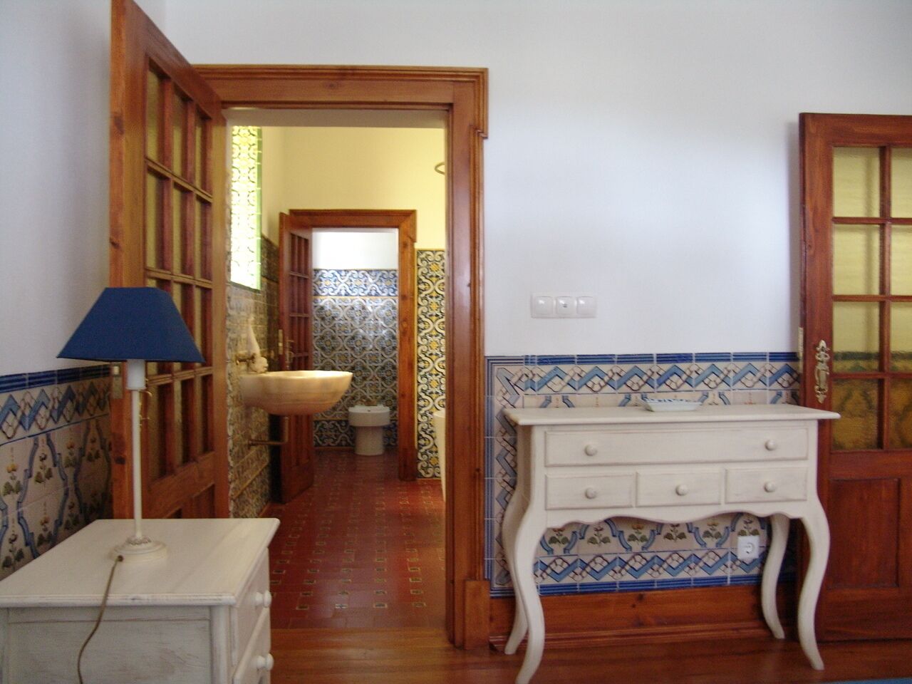 Um corredor estreito com piso de madeira, um banco branco, um abajur azul, um aparador branco e um banheiro com parede revestida em azulejos e pia.