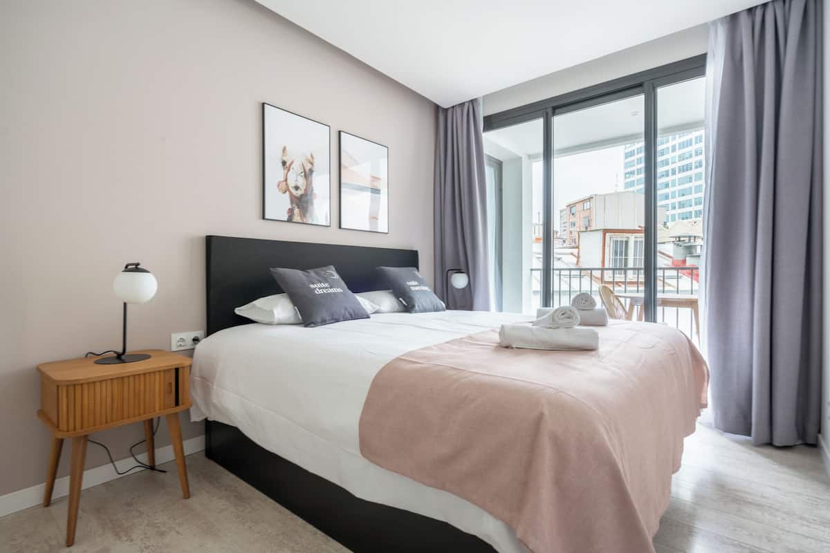 Quarto de hotel moderno com uma cama grande, uma mesa de cabeceira com abajur e vista para a cidade através de amplas janelas.