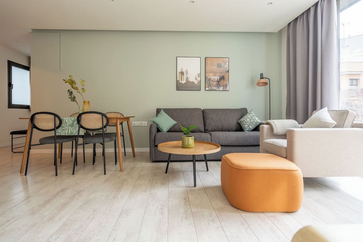 Uma sala moderna com um sofá cinzento, uma mesa de centro redonda e um espaço de jantar com cadeiras e um vaso de plantas.