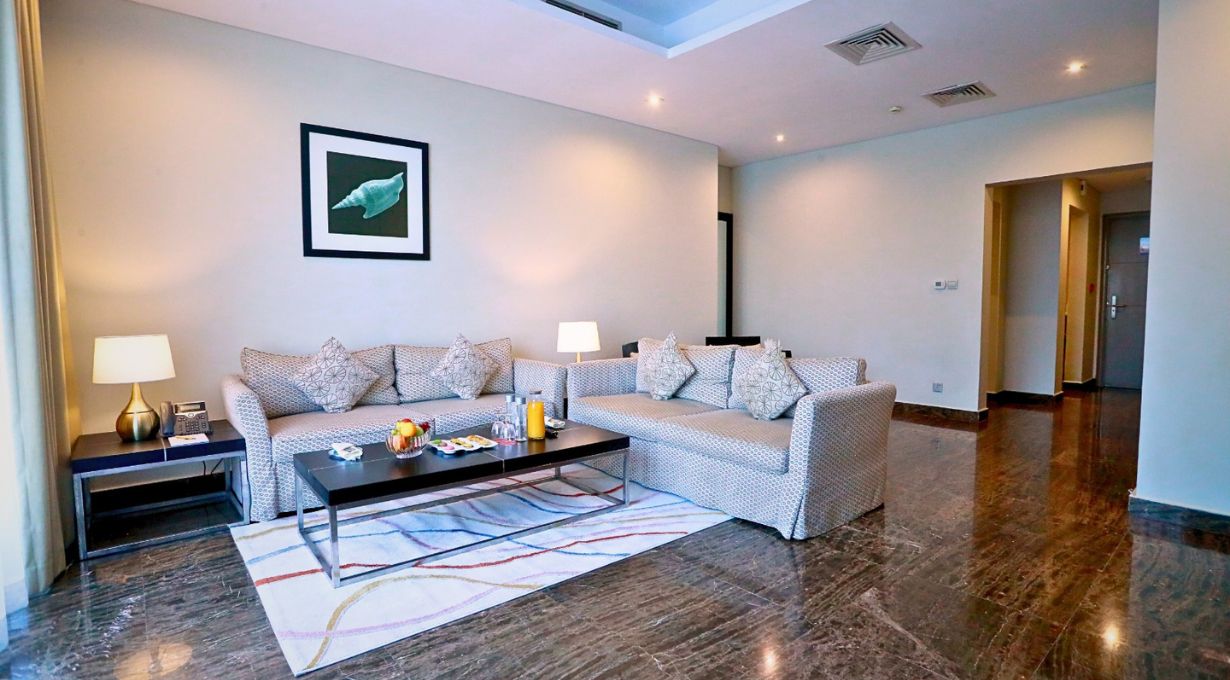 Uma sala de estar moderna com um conjunto de sofás, uma mesa de centro e um tapete. Há um quadro na parede e um abajur sobre uma mesinha lateral.
