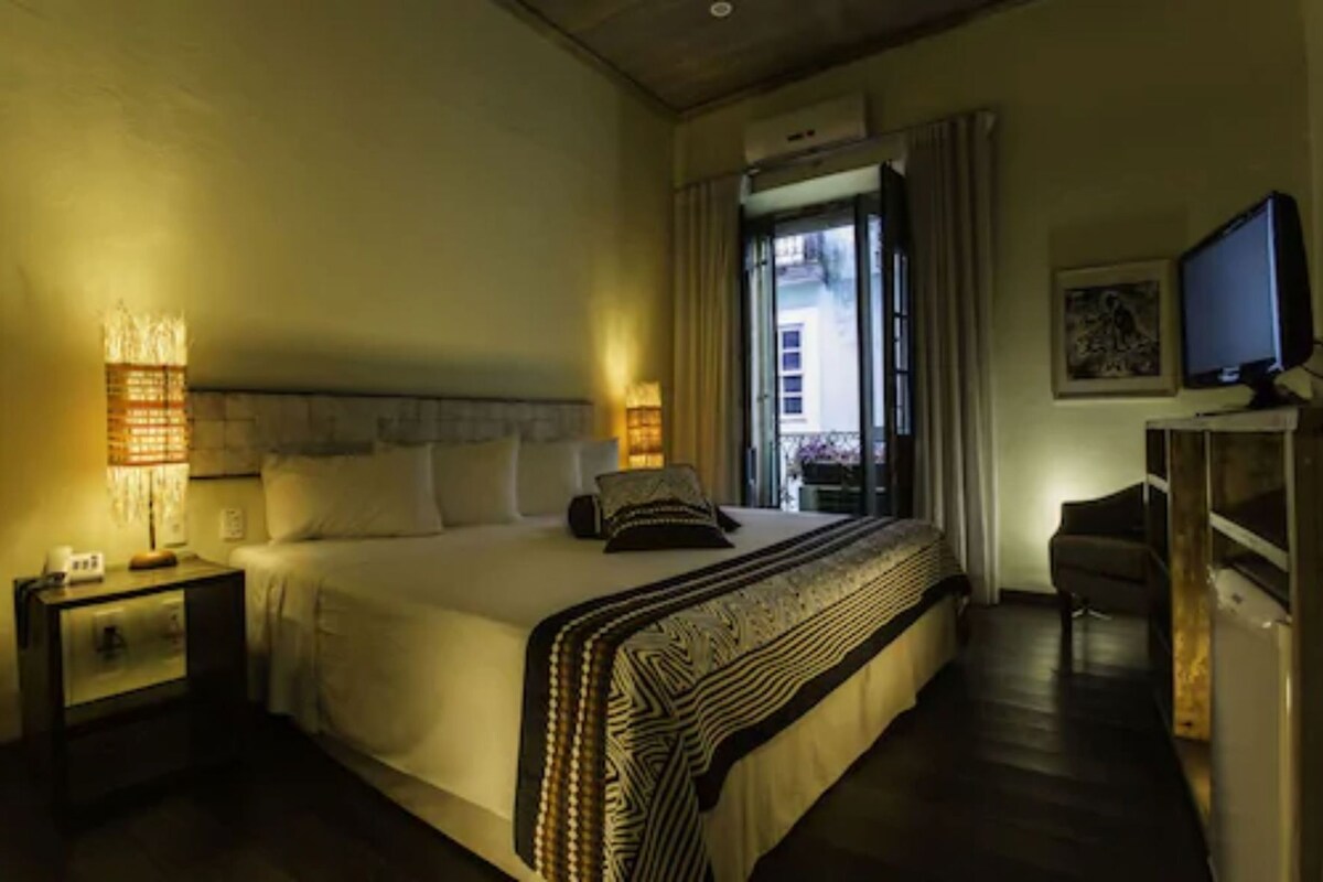 Quarto de hotel com uma cama grande, abajures, televisão e vista para o exterior.