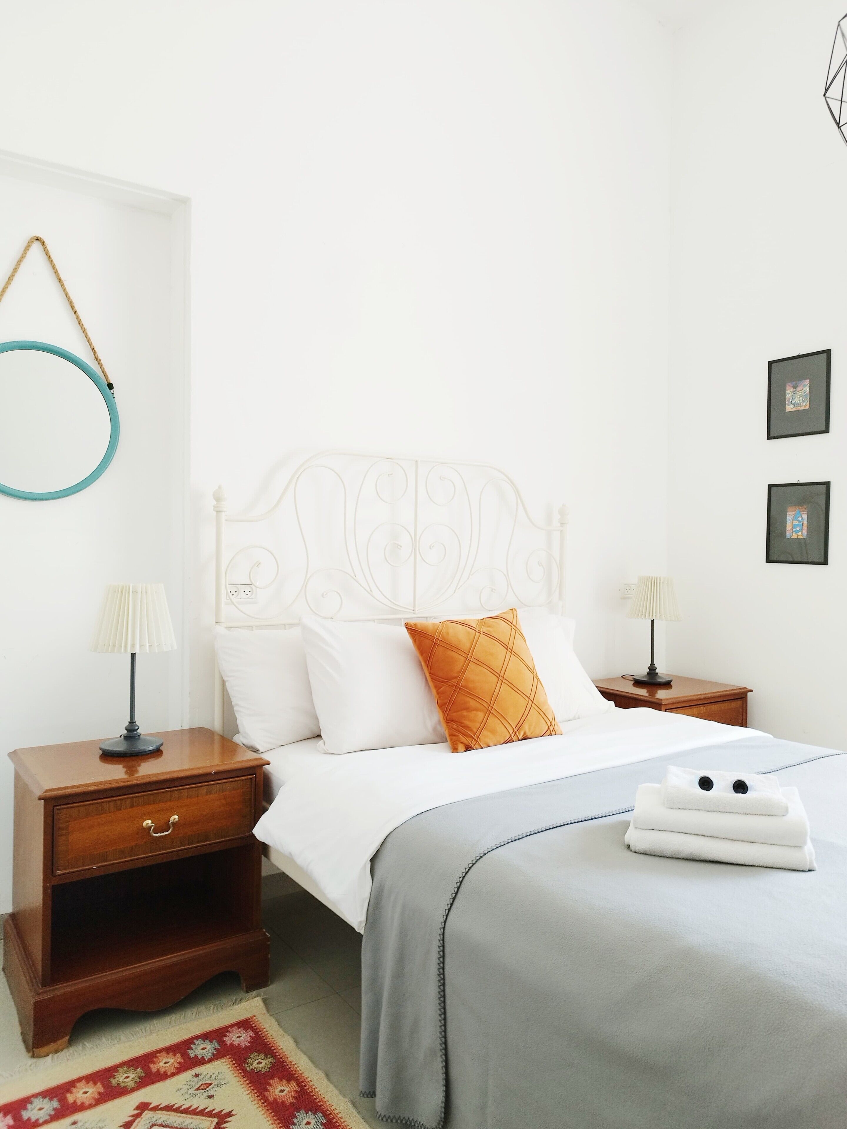 Um quarto bem organizado com uma cama, duas mesas de cabeceira, um criado-mudo, uma luminária circular na parede e um tapete estampado.