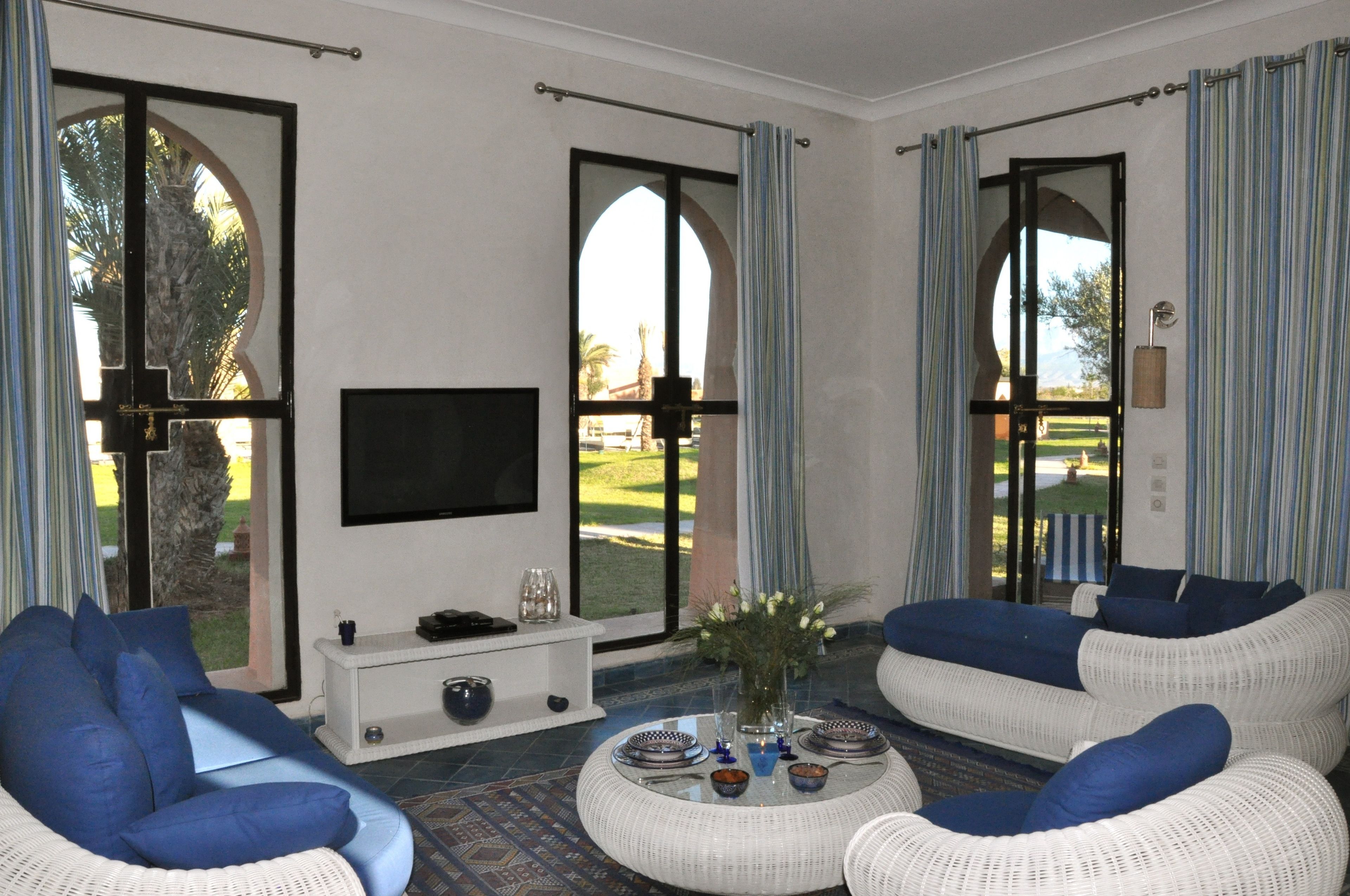 Quarto com televisão grande, móveis de vime, mesa de centro com objetos decorativos e janelas amplas com cortinas.