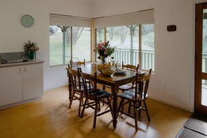 Uma sala de jantar com mesa e cadeiras de madeira, um vaso com flores e vista para o exterior através de grandes janelas.